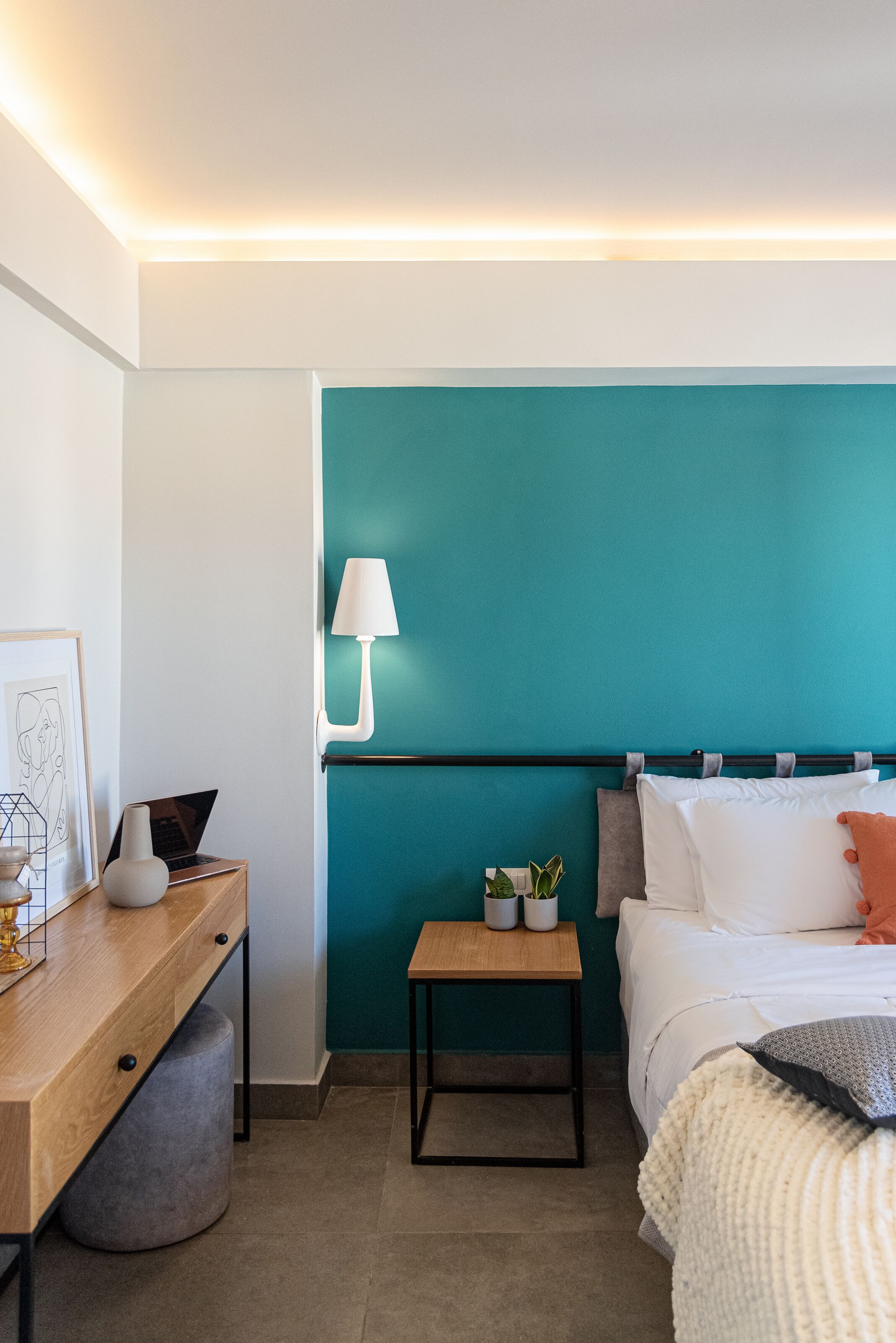 Quarto moderno com uma parede de destaque em tons de verde-água, uma escrivaninha de madeira, uma cama com roupa de cama branca e uma pequena mesa de cabeceira com uma luminária.