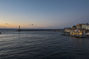 Uma cidade costeira ao entardecer, com um farol imponente e um edifício histórico com cúpulas.