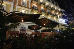 Uma área de refeições ao ar livre com guarda-sóis e mesas, um edifício com varandas e um letreiro de hotel.