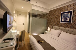 Quarto de hotel com cama, escrivaninha com televisão, cadeira, mesa de cabeceira com abajur e área de chuveiro.