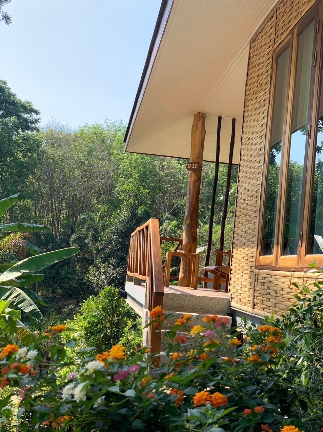 Uma casa moderna com uma grande janela, um deck de madeira e um jardim exuberante com diversas plantas e flores.