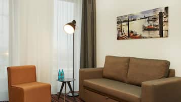 Quarto de hotel com sofá, cadeira, abajur, mesa pequena com garrafas e uma pintura de uma cena portuária.