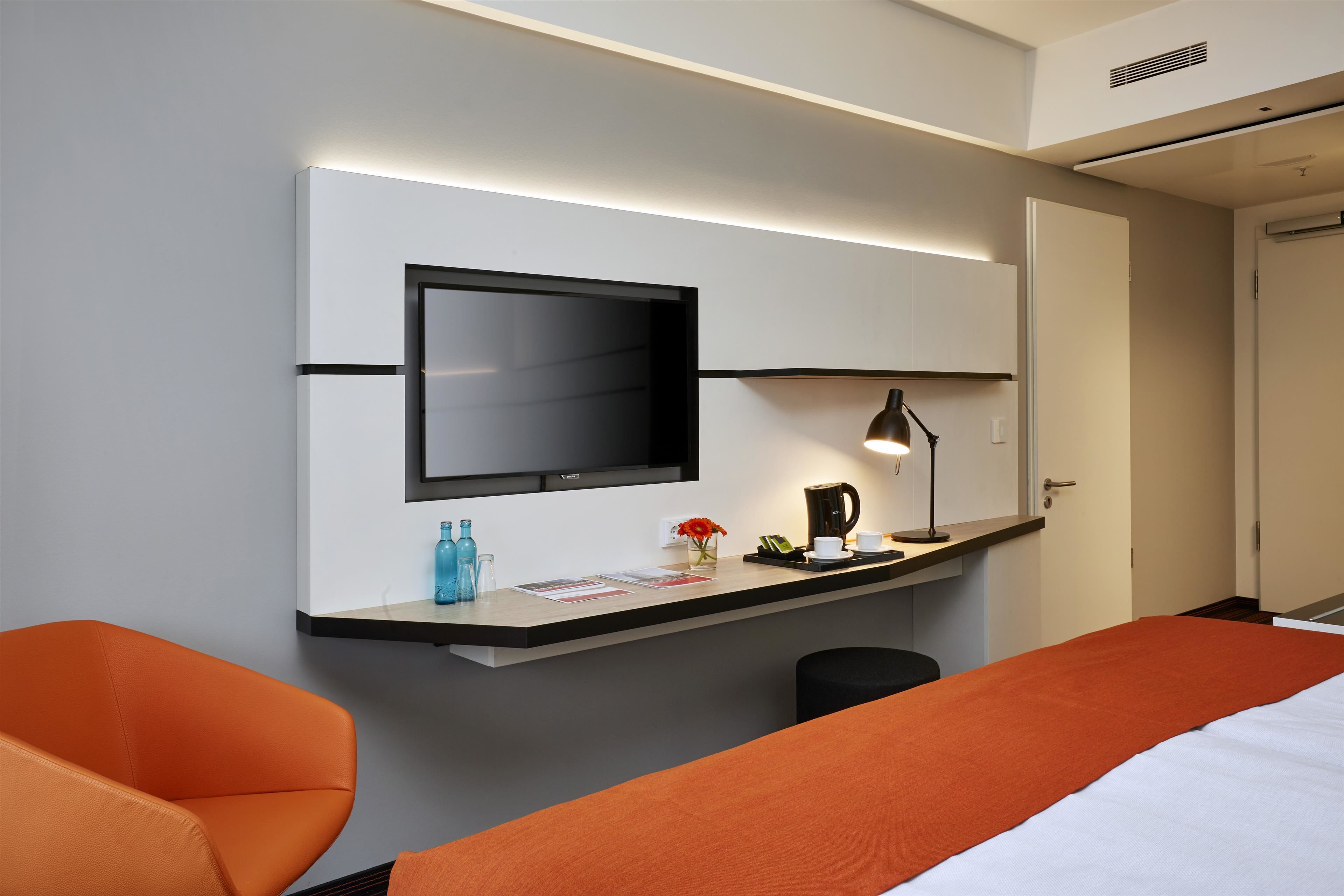 Quarto de hotel com cama, escrivaninha, televisão de tela plana, luminária e pequena mesa com cafeteira.