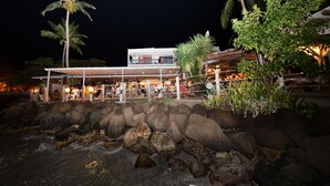 Restaurante à beira-mar com área de estar coberta ao ar livre, palmeiras e costa rochosa.