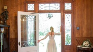 Interior de madeira com um grande candelabro, uma porta com vitrais e uma pessoa de vestido branco.