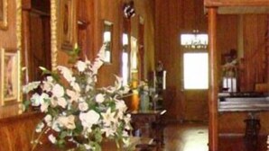 Interior de madeira com um candelabro, uma mesa com flores e uma janela com luz natural.