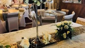 Um salão de festas com mesas redondas cobertas por toalhas brancas, decorado com arranjos florais e um caminho de mesa com franjas douradas.