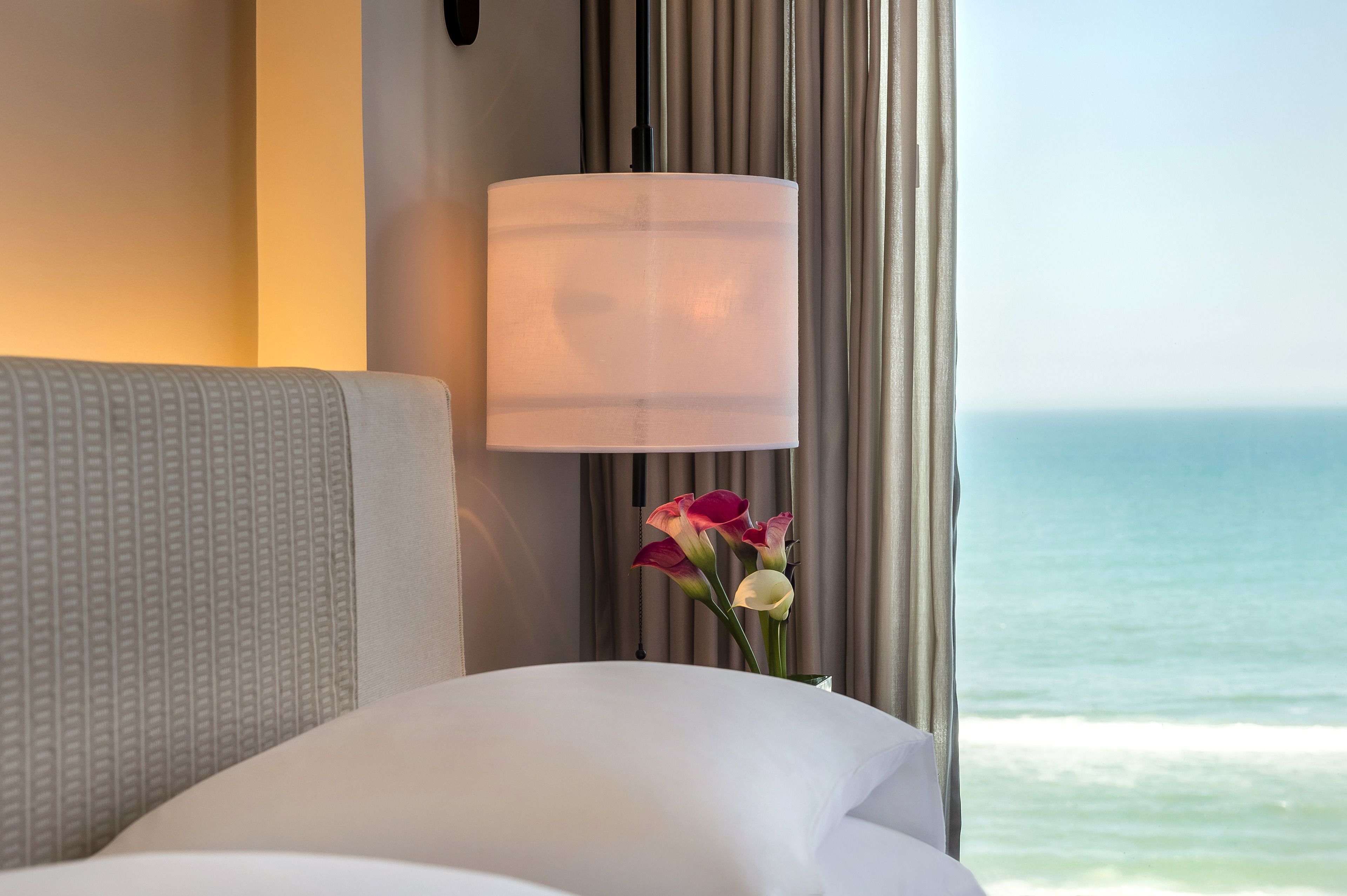 Quarto de hotel com cama, abajur e vista para o mar.