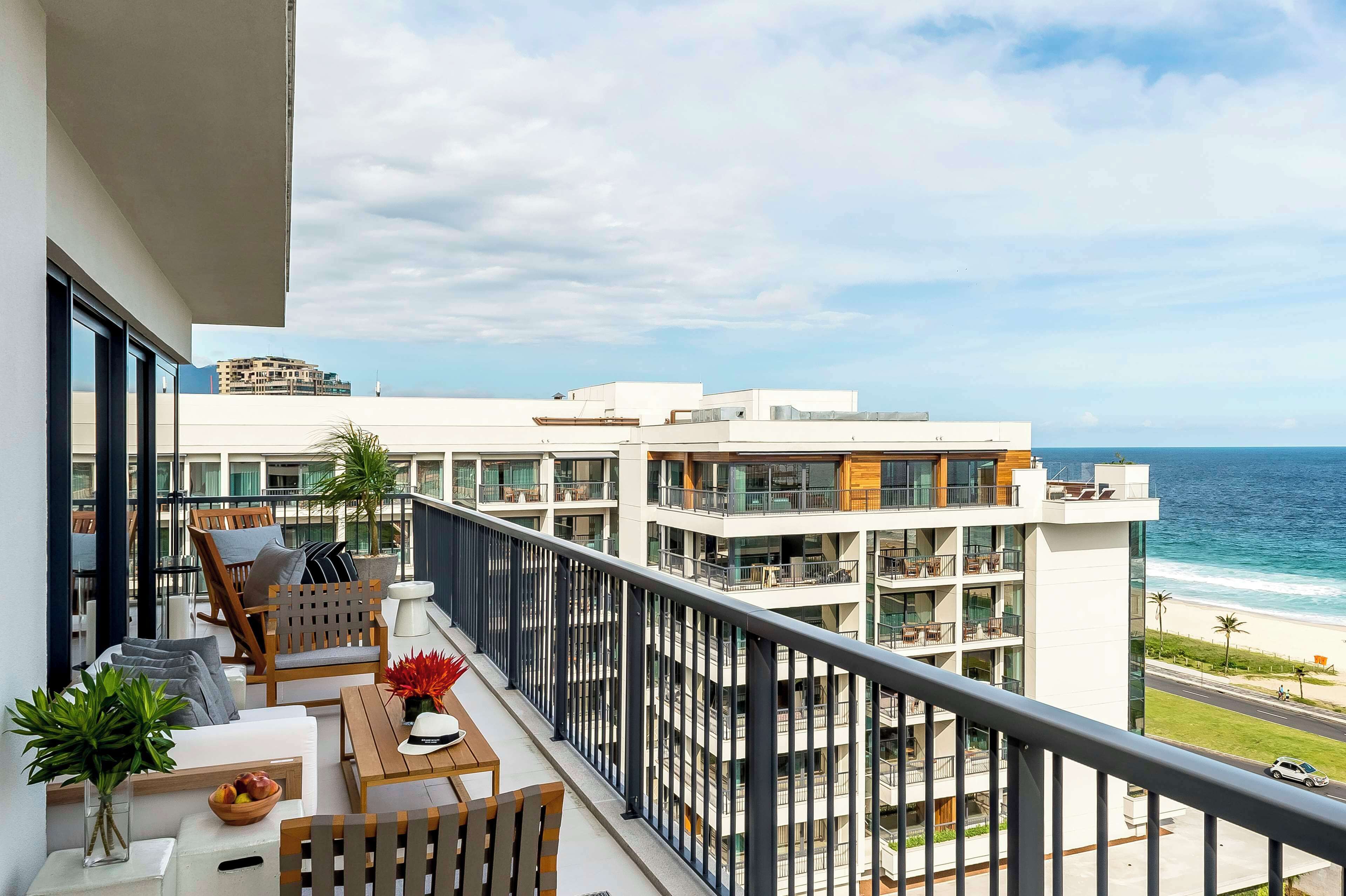 Uma varanda com mobiliário externo, vista para o oceano e um edifício de vários andares ao fundo.