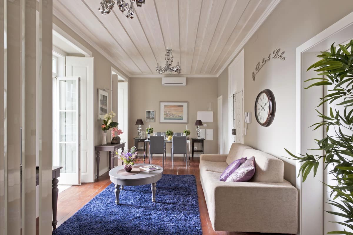 Uma sala de estar com um sofá bege, um tapete azul, uma área de jantar com mesa e cadeiras, e uma cozinha ao fundo.
