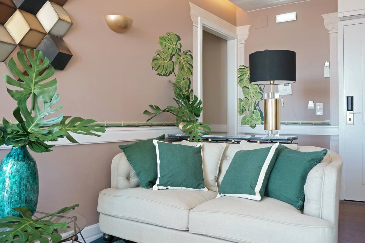 Uma sala de estar com um sofá bege, almofadas verdes, um objeto decorativo na parede e um abajur.