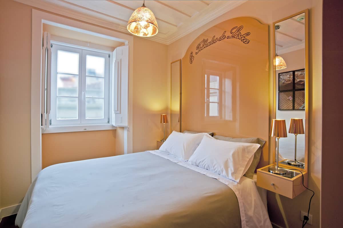 Quarto de hotel com cama, mesa de cabeceira, luminária, espelho e janela com persianas.