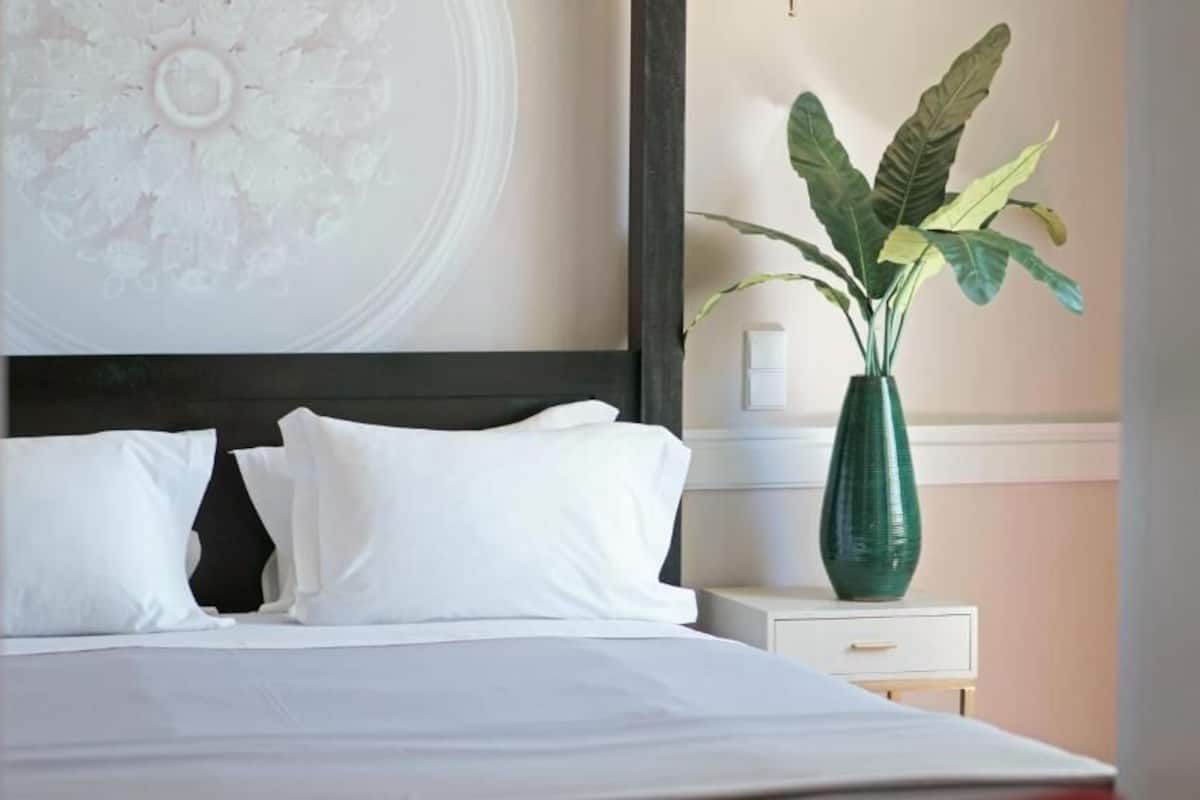Cama bem arrumada com lençóis brancos, cabeceira escura e planta em vaso sobre uma mesa de cabeceira.