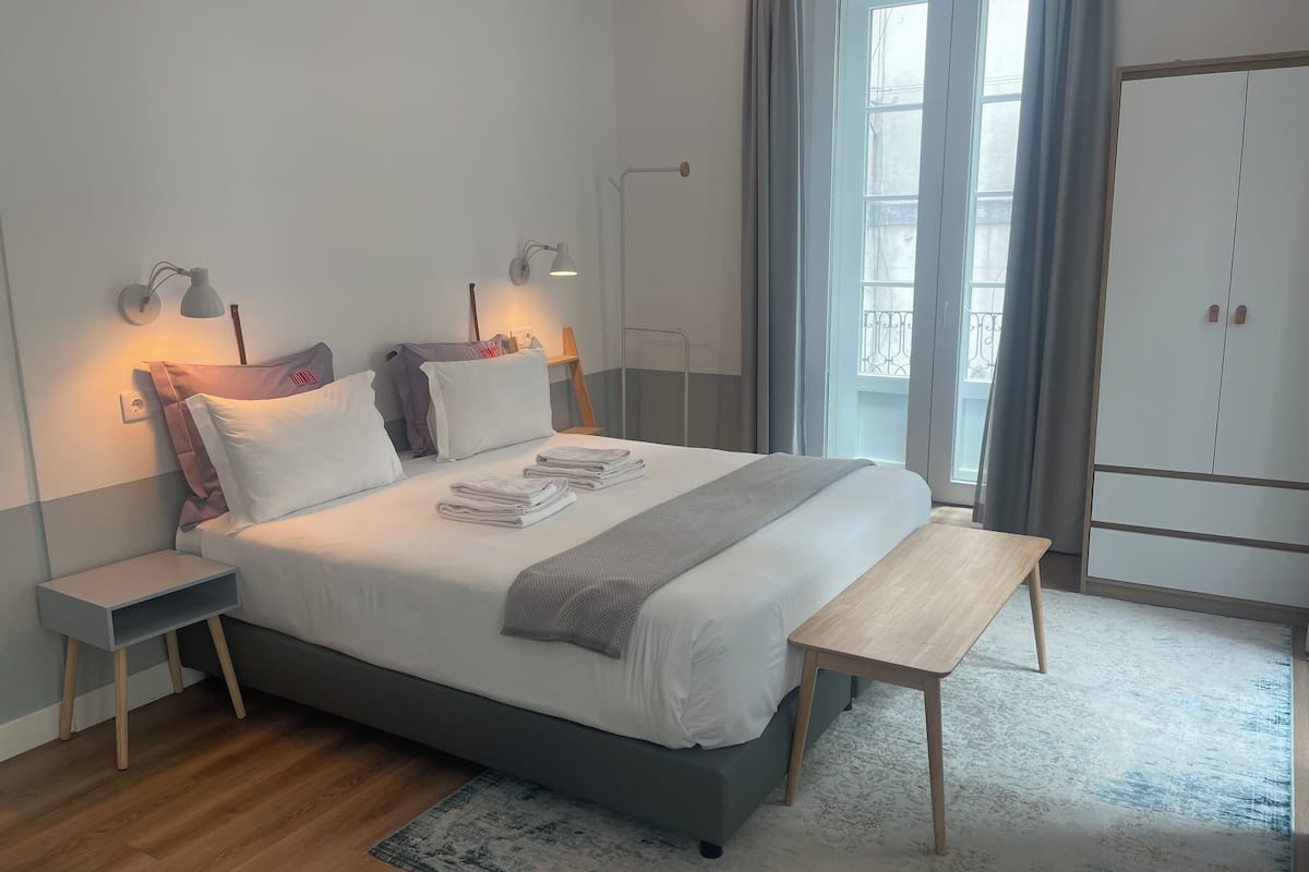 Quarto moderno com cama, mesas de cabeceira, um banco, guarda-roupa e vista para o exterior.