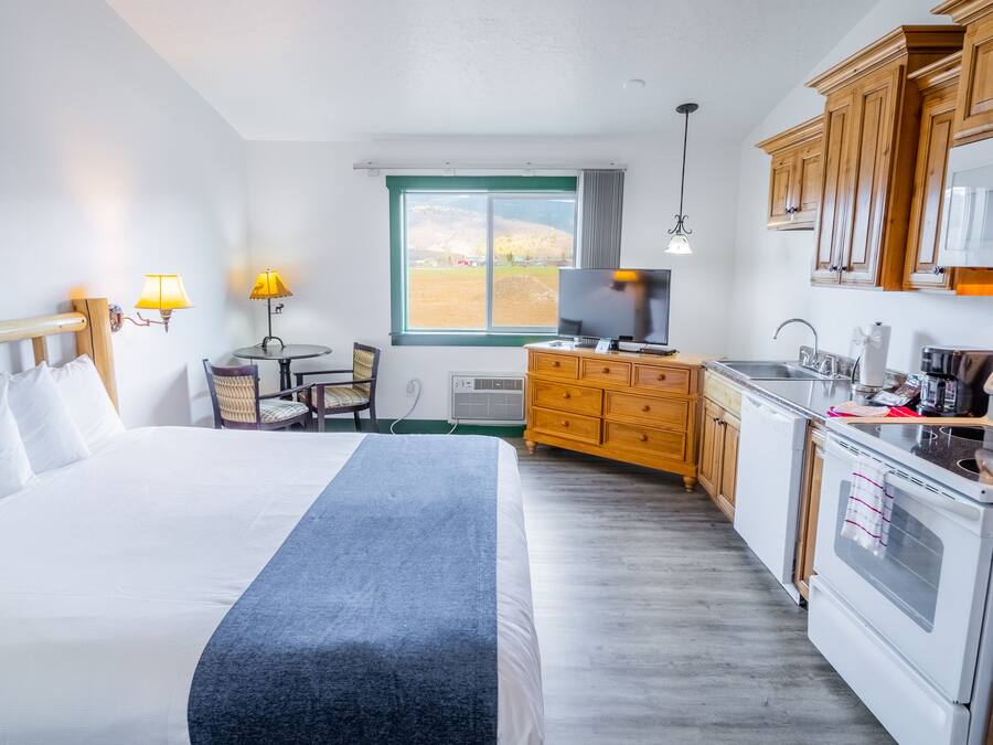 Quarto de hotel com cama, luminárias de cabeceira, TV, uma pequena mesa com cadeira, janela com vista, cozinha compacta com fogão e armários de madeira.
