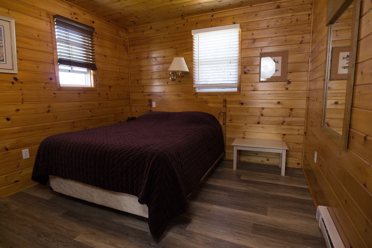 Quarto de cabana de madeira com cama, mesa de cabeceira, abajur e duas janelas.