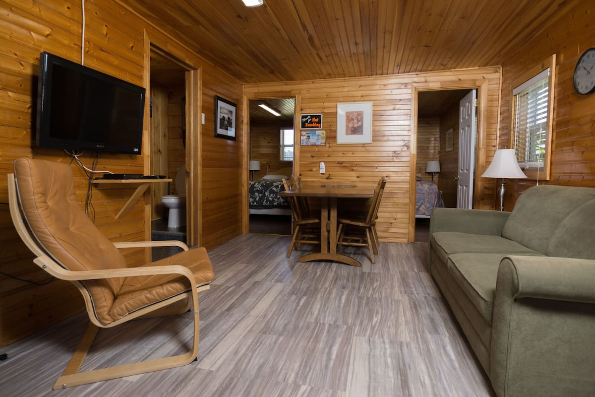 Interior de uma cabana de madeira com televisão, mesa de jantar, sofá e cadeira de balanço.