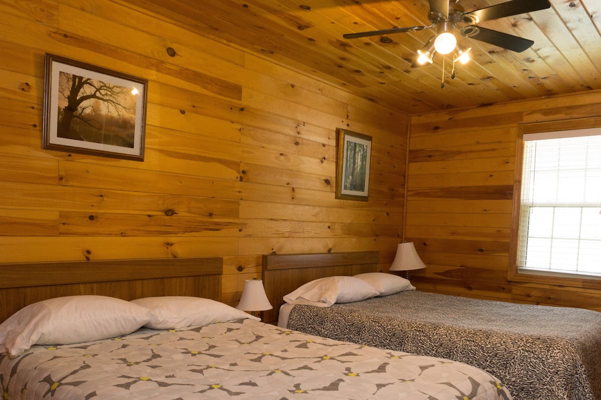 Quarto de cabana de madeira com duas camas, um ventilador de teto e uma janela.