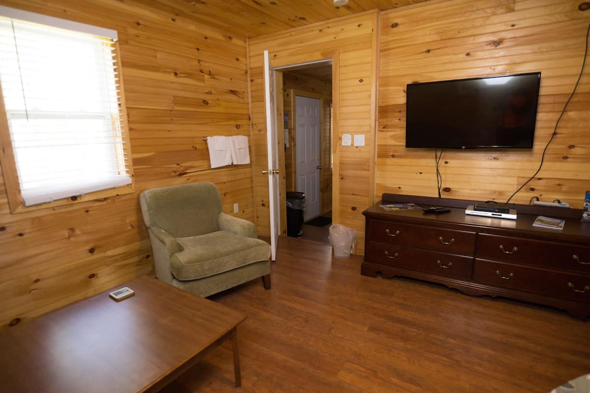 Quarto com paredes de madeira, televisão fixada na parede, cómoda de madeira, cadeira e uma pequena mesa.