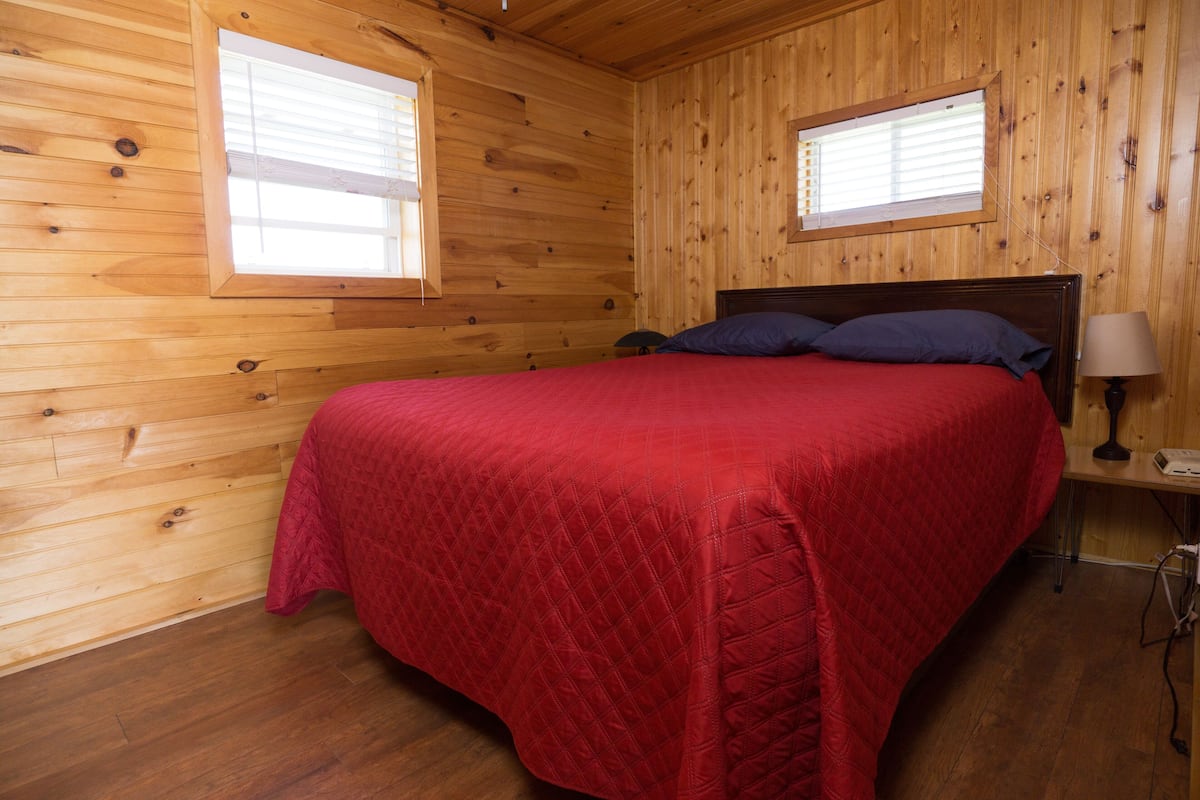 Quarto com painéis de madeira, uma cama, duas janelas e uma mesa de cabeceira com abajur.