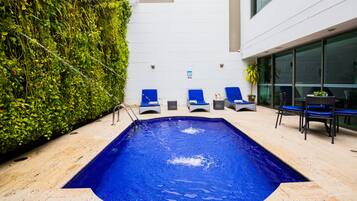 Uma piscina com água azul, rodeada por um muro branco e um viveiro verde. Há espreguiçadeiras azuis e uma mesa com cadeiras.