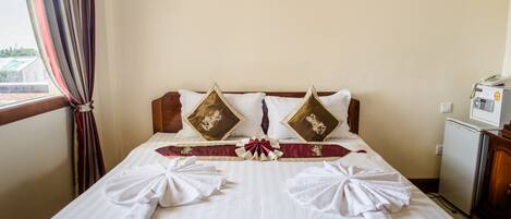Quarto de hotel com cama bem arrumada, janela com cortinas e um pequeno frigorífico.