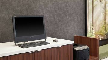 Um computador desktop Lenovo sobre uma mesa branca, com teclado e rato, uma impressora e uma janela com vista para uma floresta.