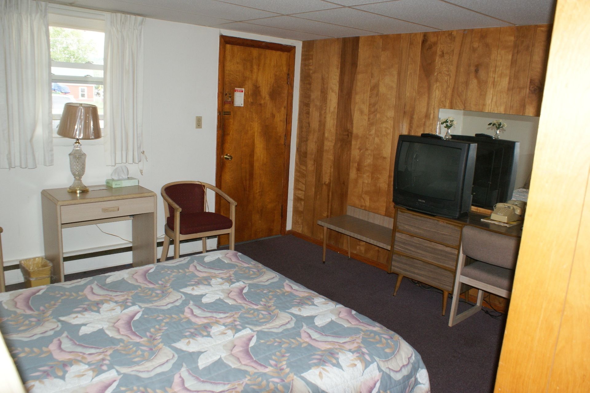Quarto de hotel com cama, escrivaninha, cadeira, televisão e parede de painel de madeira.