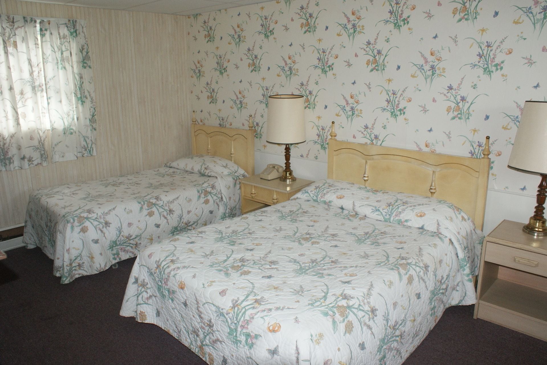 Quarto de hotel com duas camas de solteiro, roupa de cama com padrão floral, mesa de cabeceira com abajur e telefone.