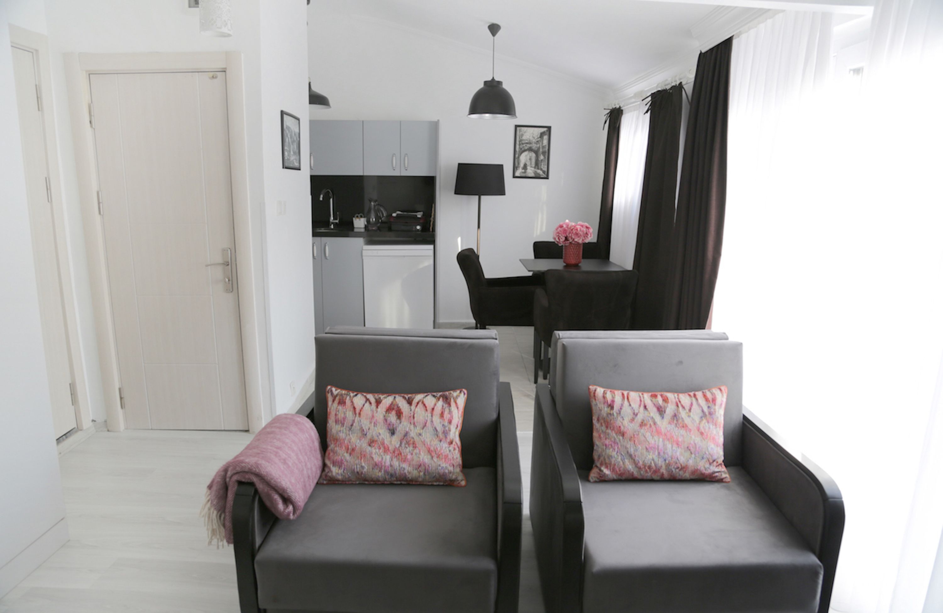 Uma sala de estar moderna com dois sofás cinzas, um cobertor rosa, uma mesa de jantar com cadeiras, uma área de cozinha e uma grande janela com cortinas.