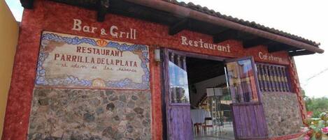 Um restaurante com fachada de pedra e portas abertas, exibindo uma placa que diz "Bar & Grill Restaurant Parrilla de la Plata".