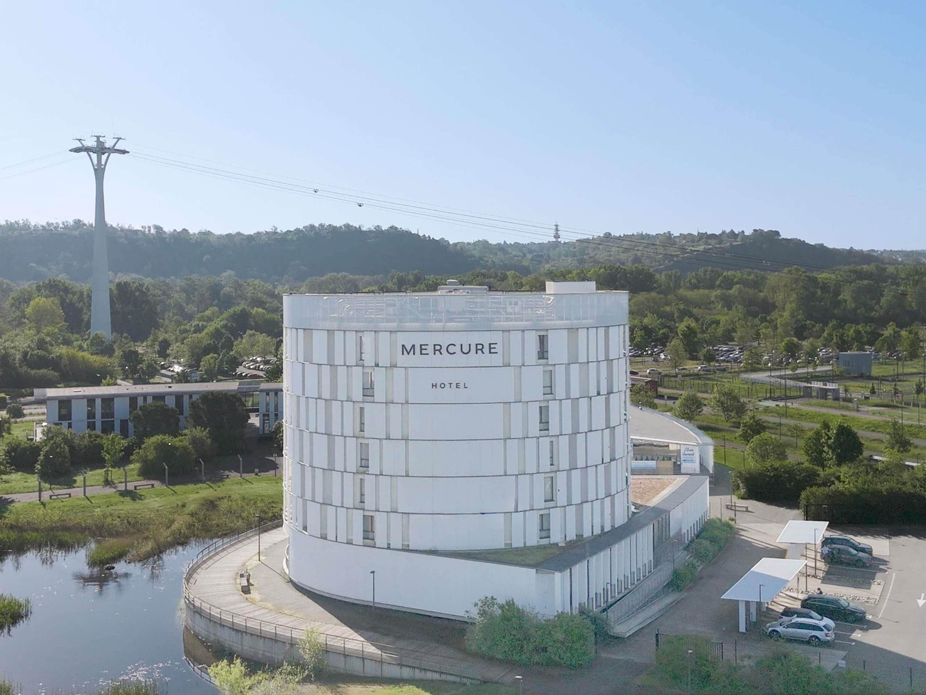 Um edifício moderno de hotel com formato cilíndrico chamado 'Mercure', com telhado plano, situado perto de um corpo de água e rodeado de vegetação.