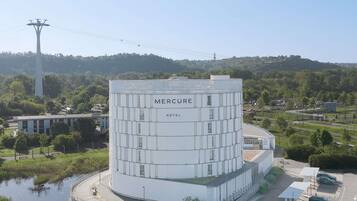Um edifício moderno de hotel com formato cilíndrico chamado 'Mercure', com telhado plano, situado perto de um corpo de água e rodeado de vegetação.