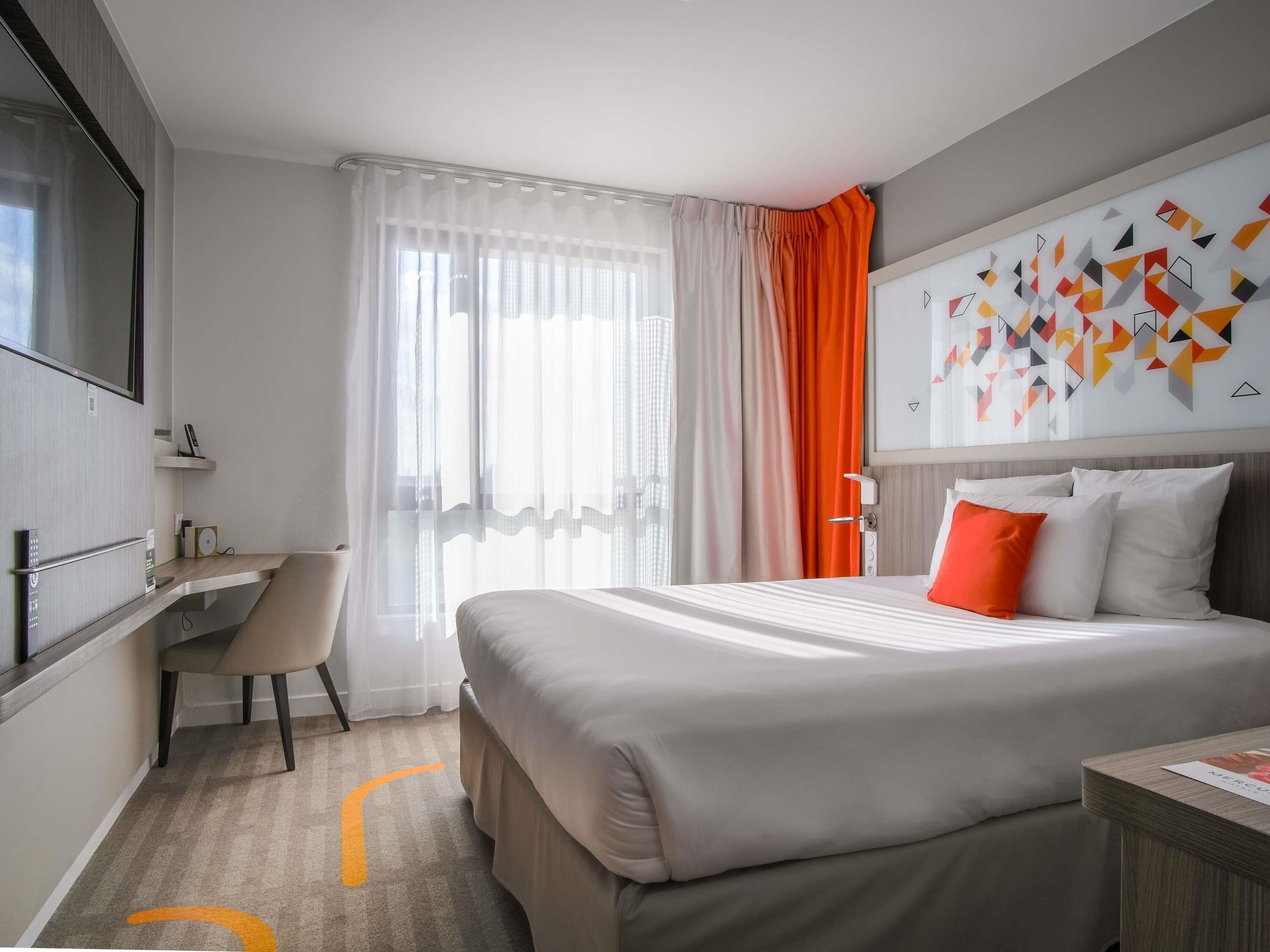 Quarto de hotel com cama, escrivaninha, cadeira, televisão e janela com cortinas.