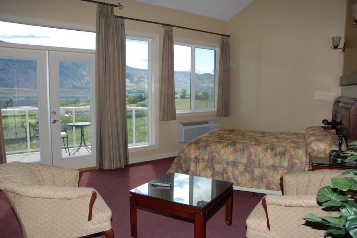 Quarto com cama, duas poltronas, mesa com tampo de vidro e vista para uma paisagem montanhosa através de grandes janelas.
