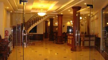 Um saguão de hotel com uma escadaria, colunas de madeira e um balcão de recepção.