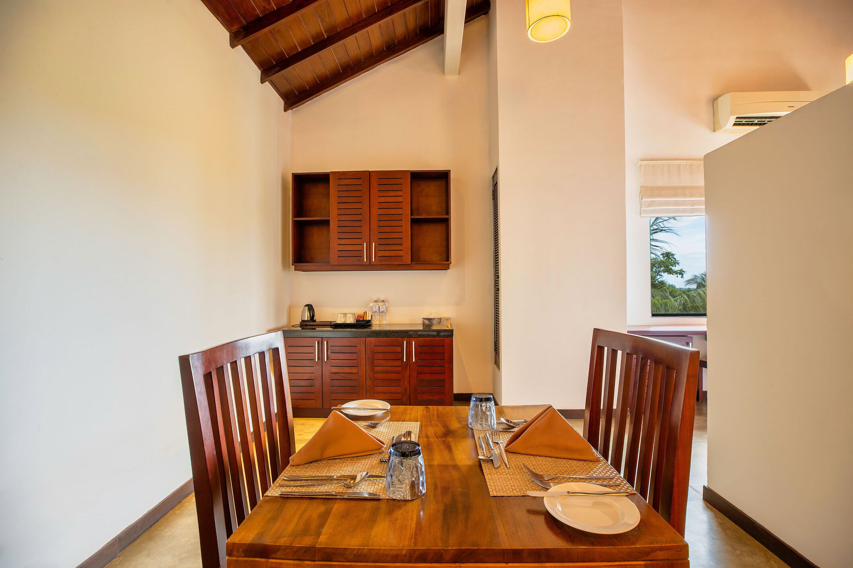 Uma sala de jantar com uma mesa de madeira posta para dois, uma cozinha ao fundo e vista para o exterior.