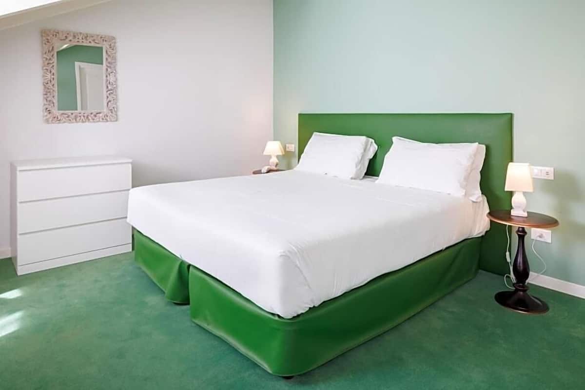 Quarto com cama estofada em tom verde, roupa de cama branca, cômoda branca e duas luminárias de cabeceira.