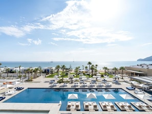 Área de piscina com espreguiçadeiras e guarda-sóis, vista para o mar e um edifício com um mural.
