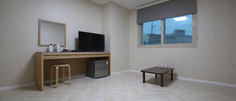Quarto com escrivaninha de madeira, televisão, mesa pequena e janela com vista para edifícios.