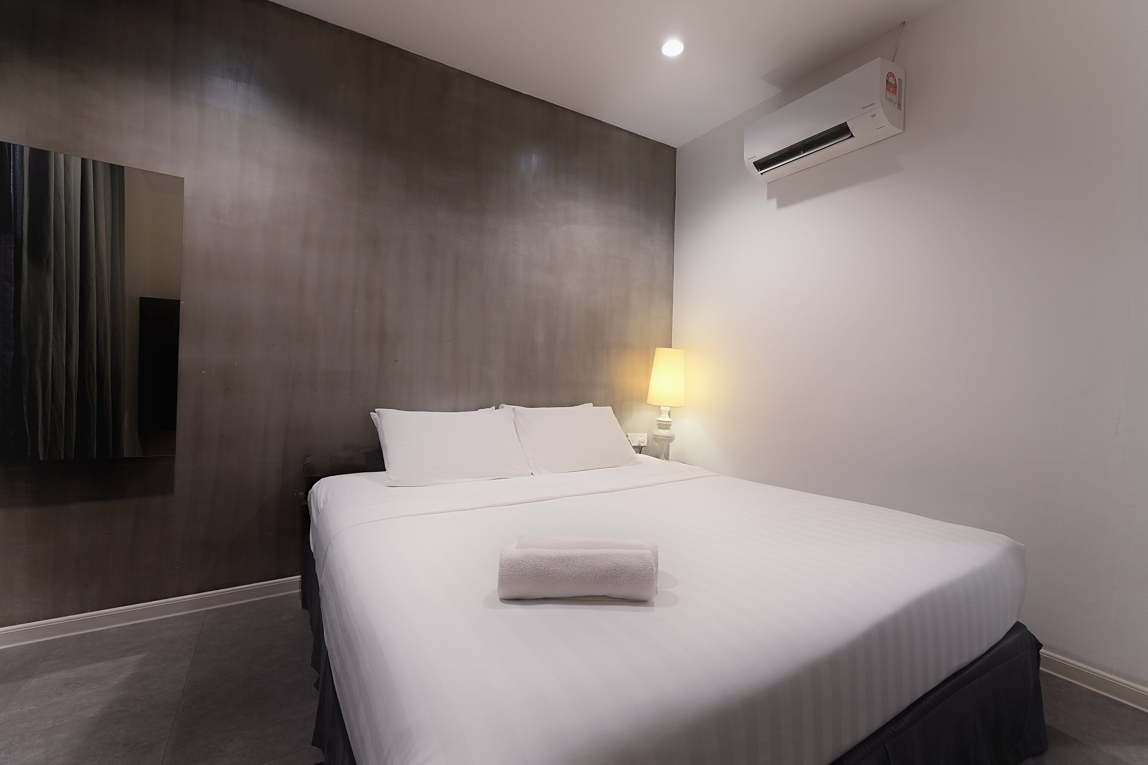 Uma cama bem arrumada com lençóis brancos e uma toalha dobrada, uma luminária de cabeceira e um ar condicionado fixado na parede em um quarto de hotel moderno.