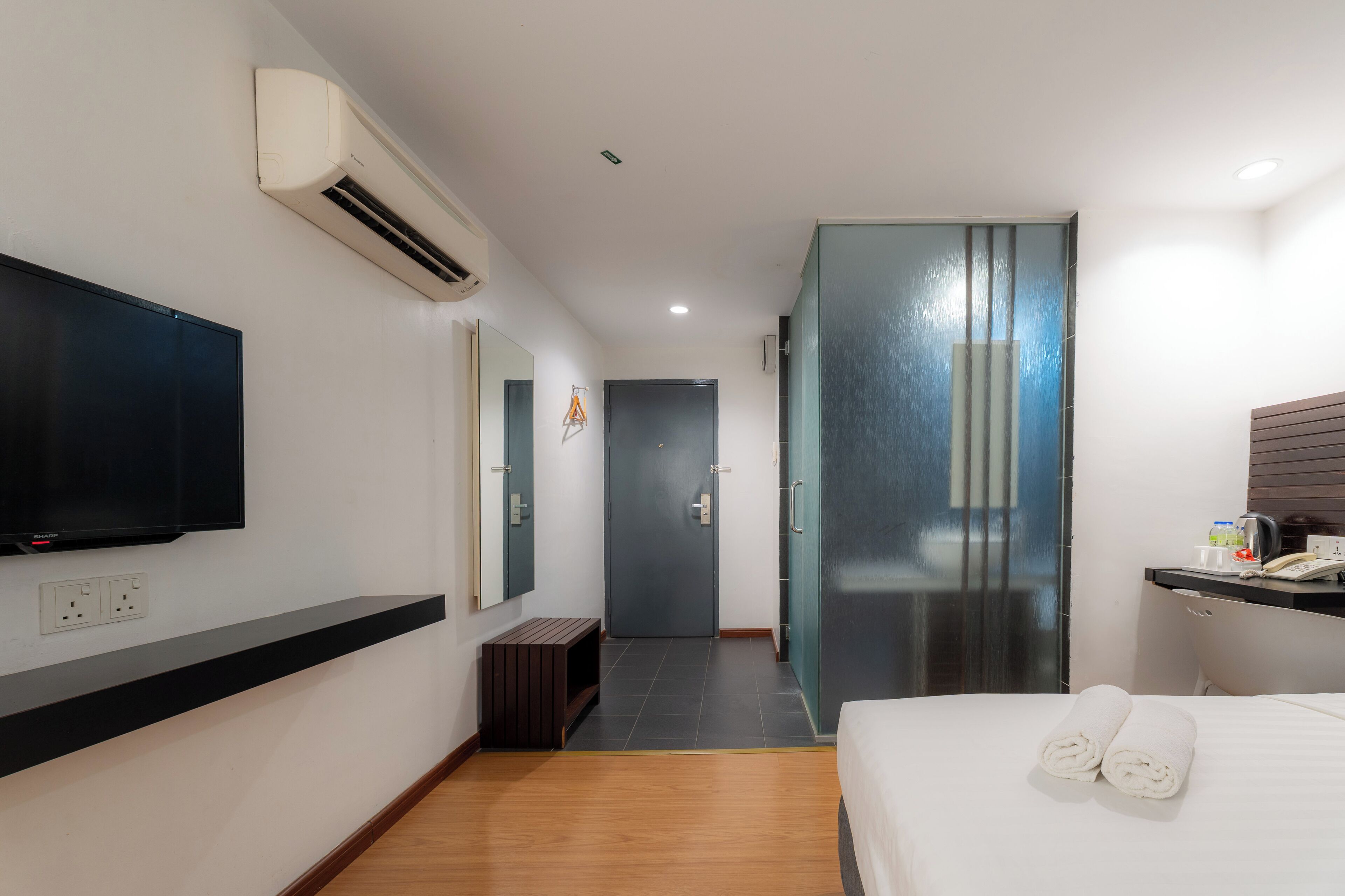 Quarto de hotel com televisão de tela plana, um espelho, uma cama com roupa de cama branca, mesa de cabeceira com telefone e uma xícara, e uma porta de correr que dá para outro cômodo.