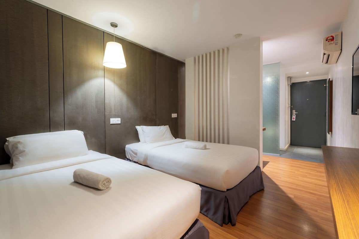 Quarto de hotel com duas camas, piso de madeira, luminária moderna e porta que dá para outro cômodo.