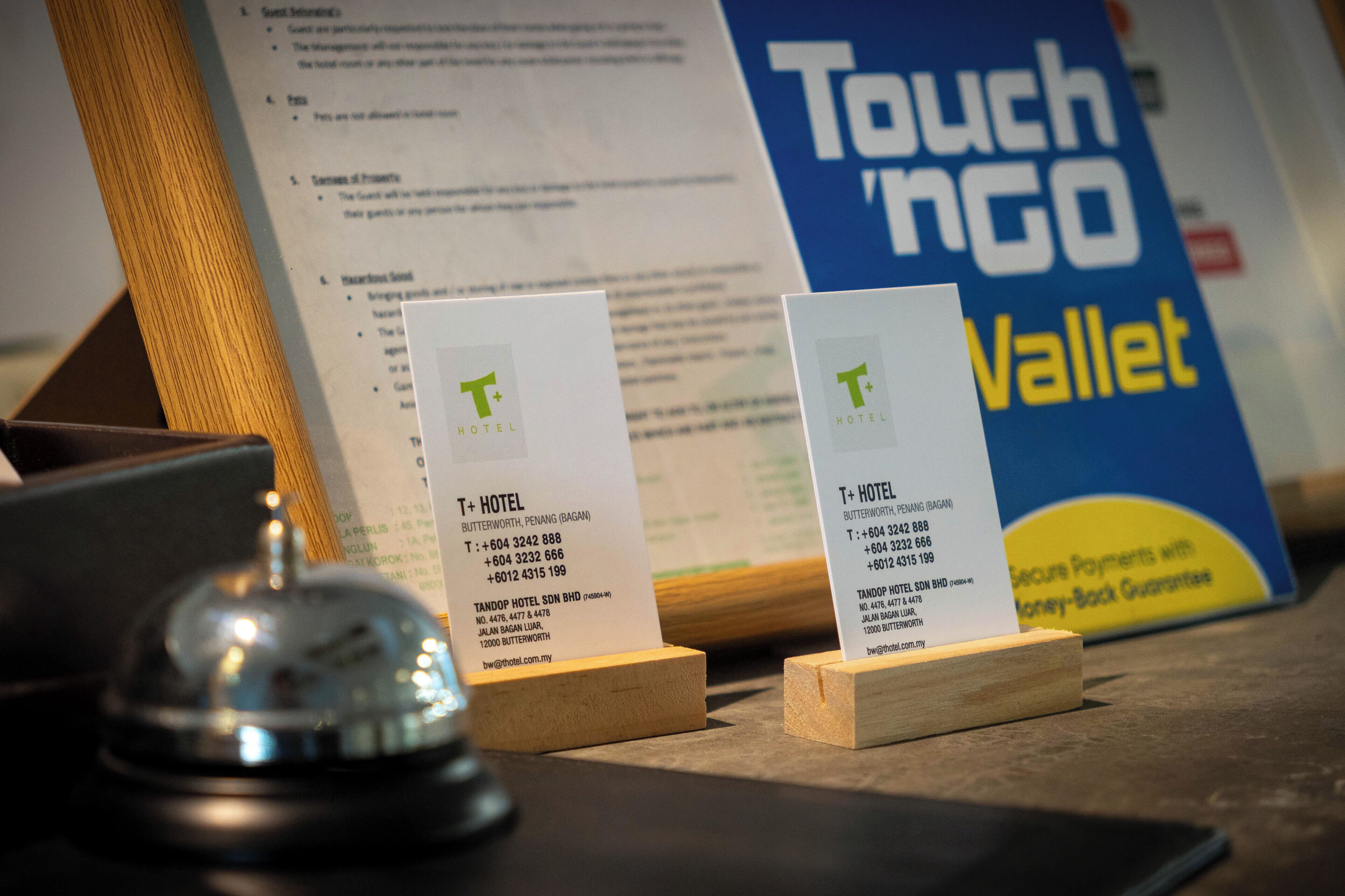 Dois cartões informativos do T+ Hotel em suportes de madeira, com um letreiro de 'Touch 'N Go Wallet' ao fundo.