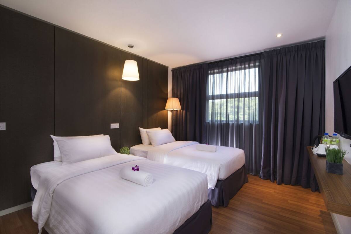 Quarto de hotel com duas camas, uma escrivaninha, uma luminária e uma janela com cortinas.