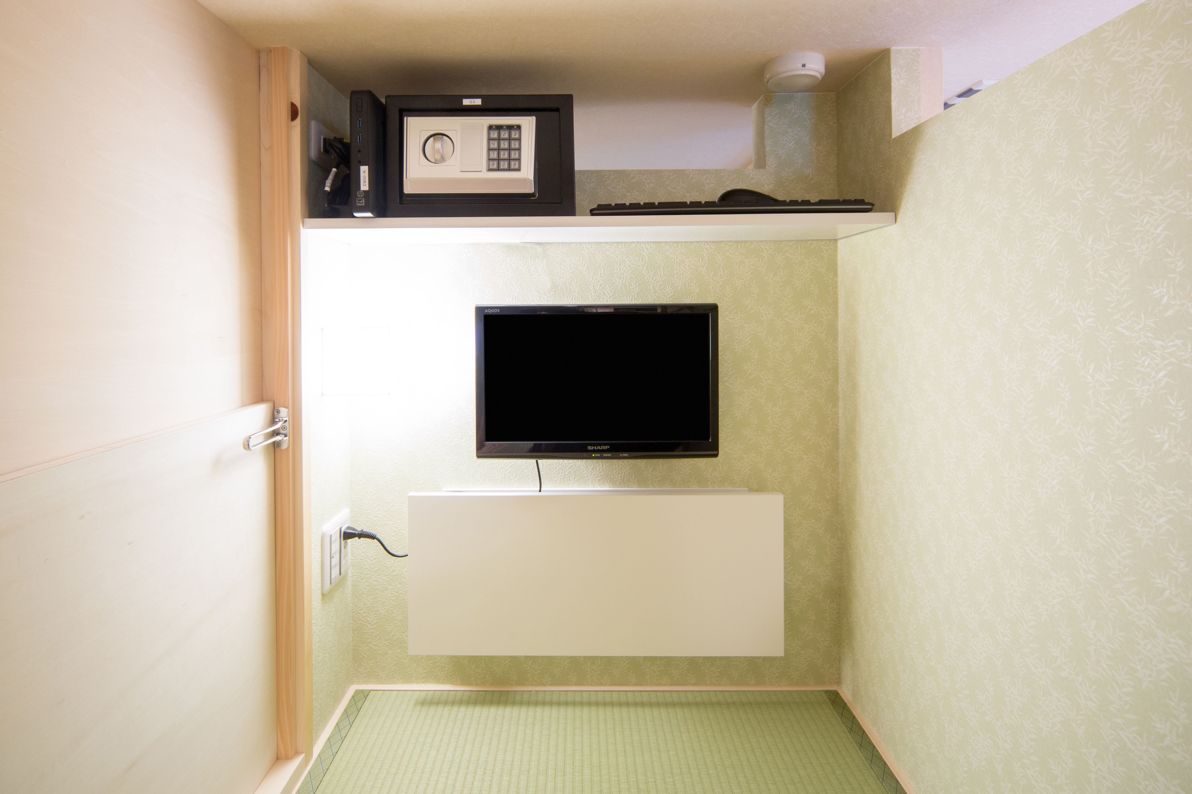 Um quarto pequeno com uma televisão presa na parede, um cofre e um armário abaixo da TV.