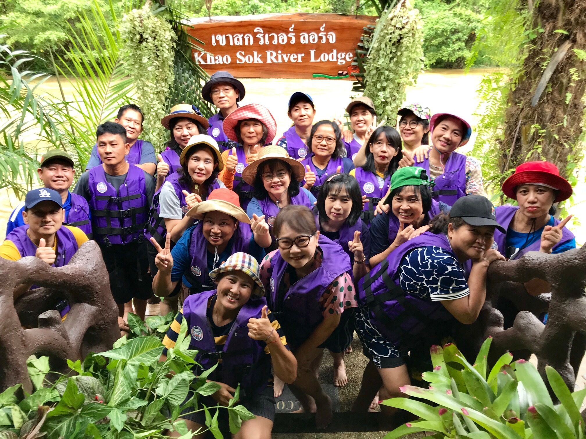 Um grupo de pessoas em coletes roxos reunido em frente a uma placa com a inscrição 'Khao Sok River Lodge'.