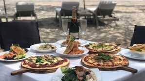 Uma mesa posta na praia com diversos pratos, incluindo pizzas e frutos do mar, e uma garrafa de champanhe.