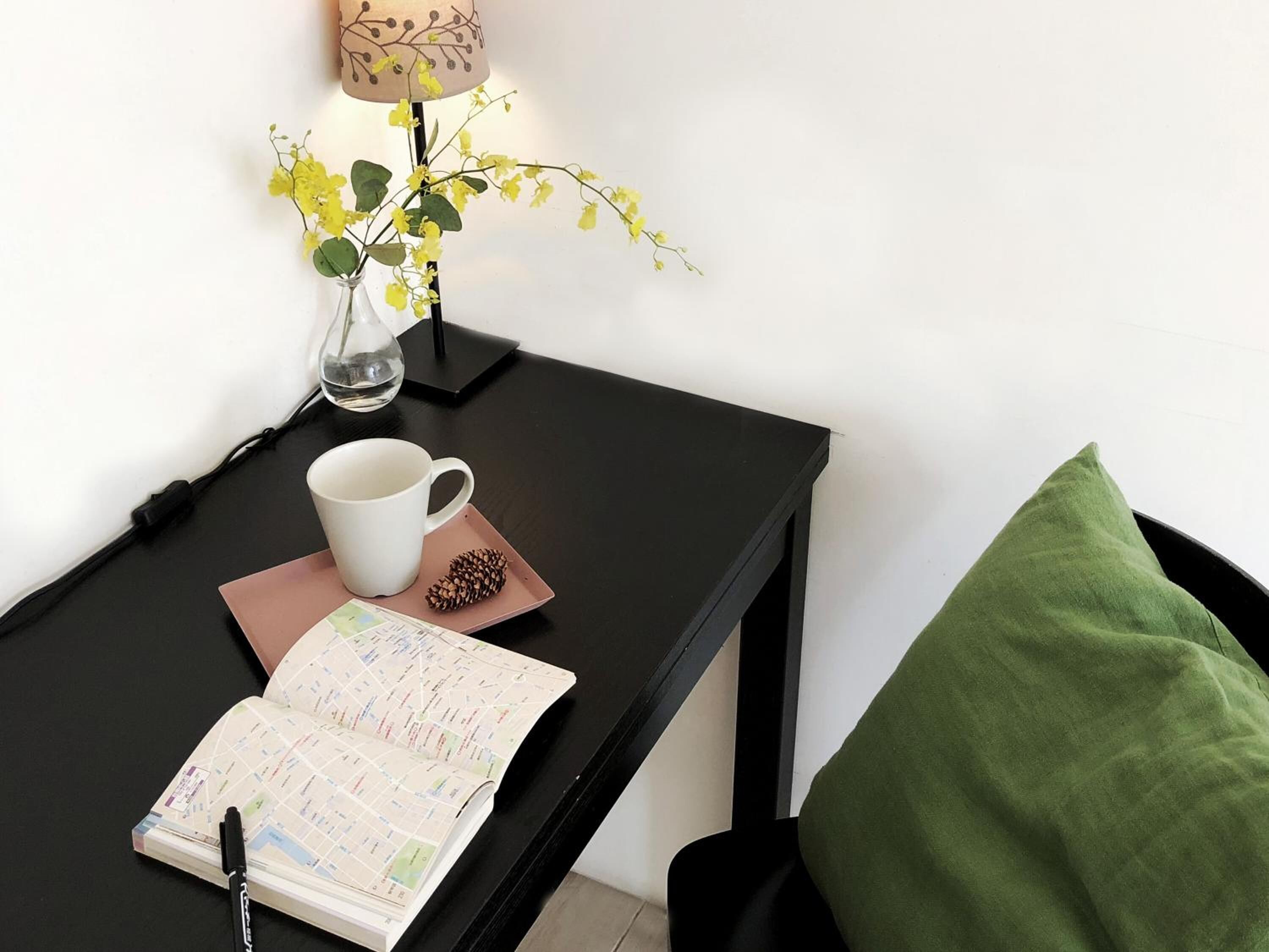Uma pequena mesa preta com uma caneca branca, um caderno, uma caneta, um pequeno vaso com flores amarelas e uma almofada verde.