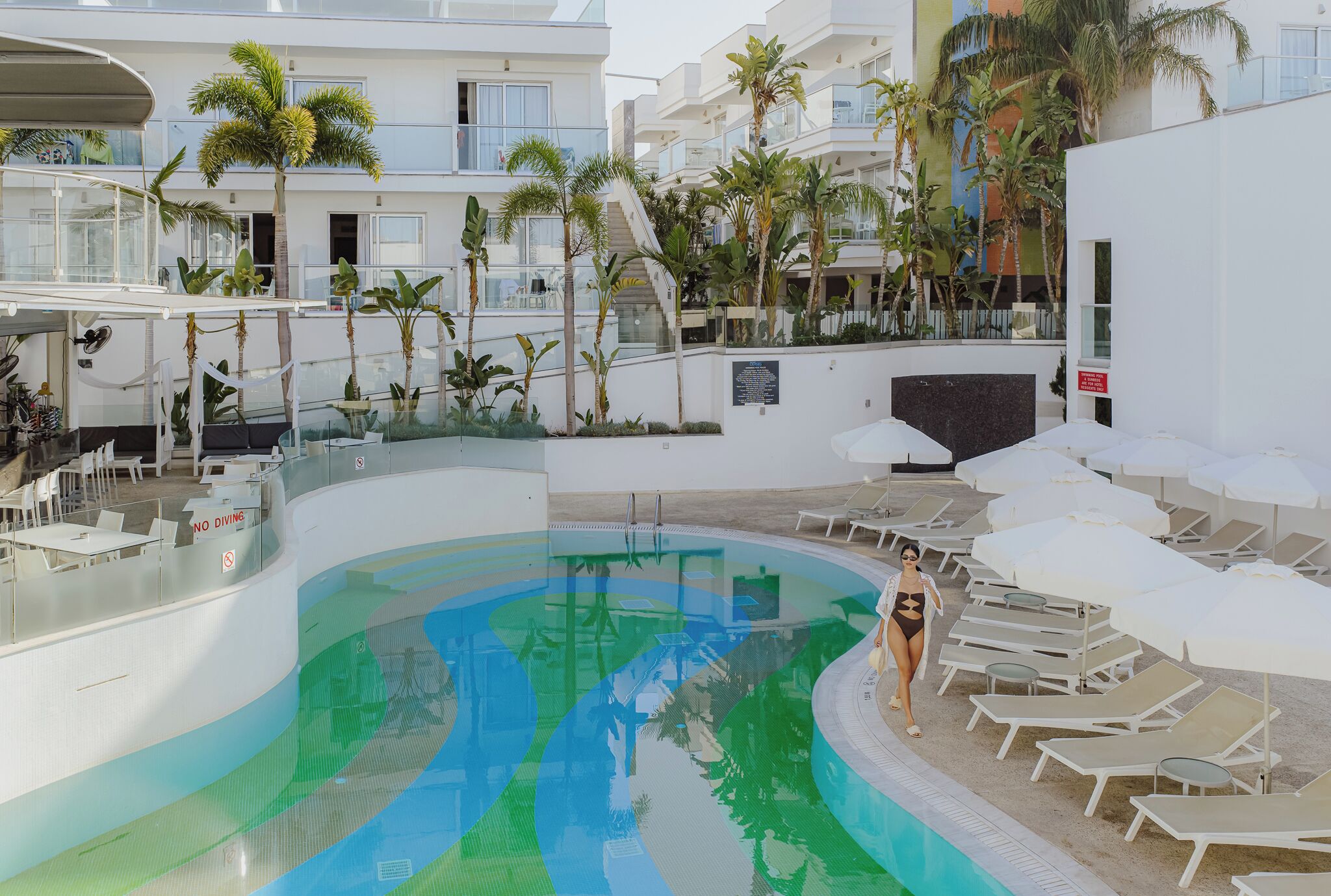 Piscina com espreguiçadeiras e guarda-sóis, cercada por palmeiras e um edifício branco.
